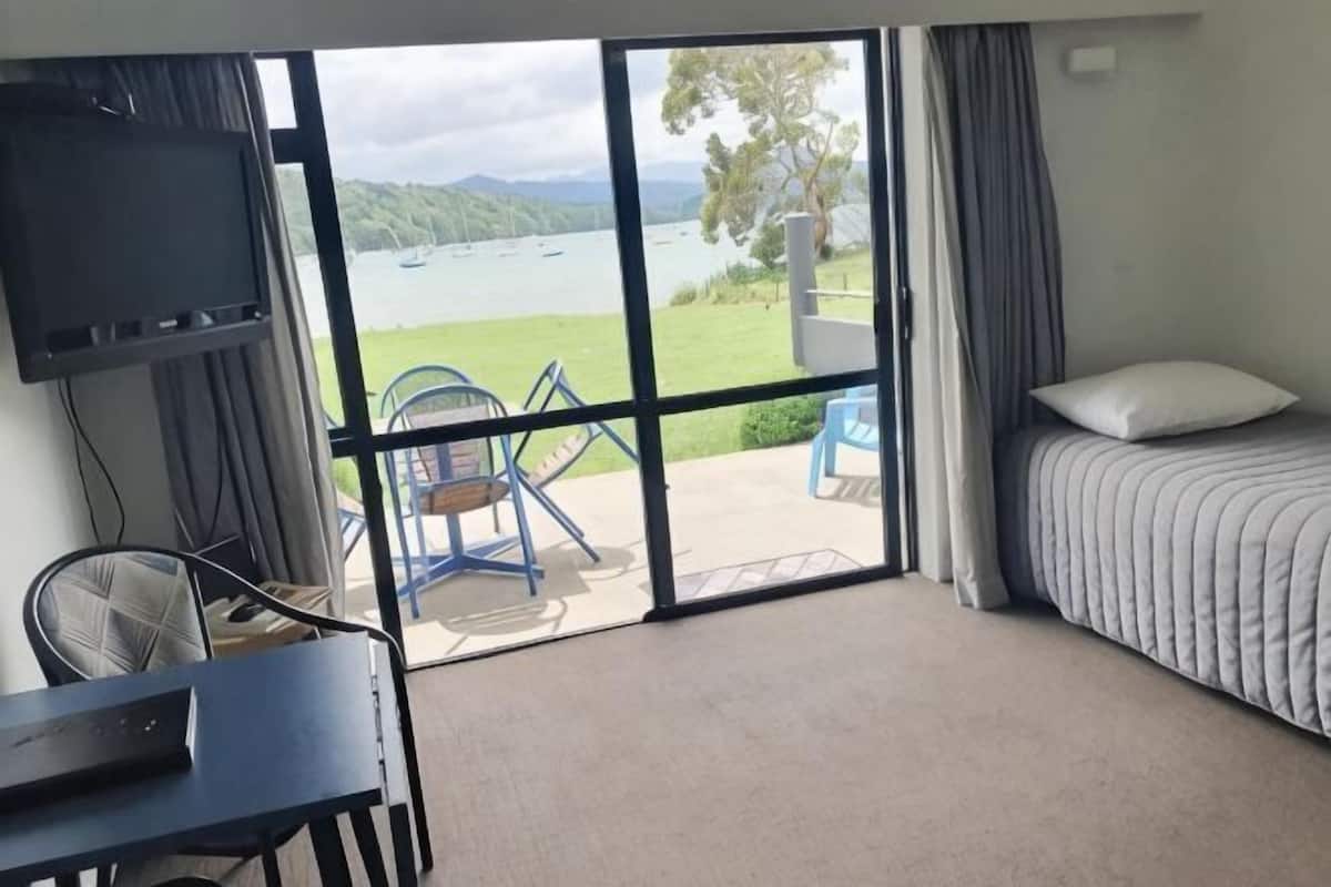 Quarto com cama, escrivaninha, cadeira, televisão e vista para um lago.