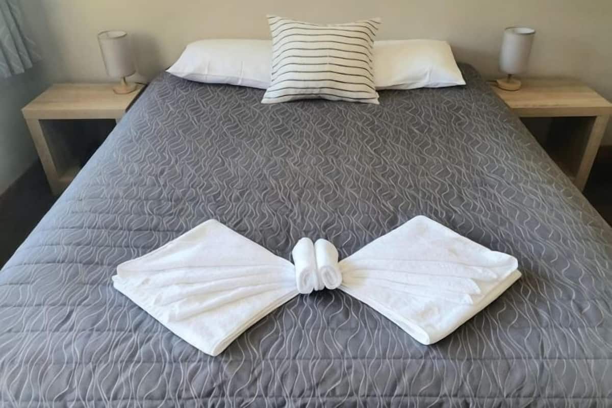 Uma cama bem arrumada com um edredom cinza, duas almofadas e uma mesa de cabeceira com abajur.