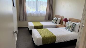 Quarto de hotel com duas camas, janela com cortinas, luminária de cabeceira e porta.