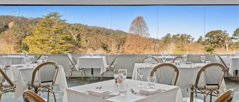 Restaurante com mesas preparadas para refeições, com vista para uma paisagem pitoresca de árvores e colinas.