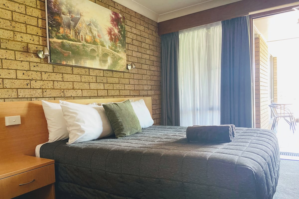 Quarto de hotel com parede de tijolos, uma pintura, cama com almofadas, mesa de cabeceira de madeira e vista para o exterior.