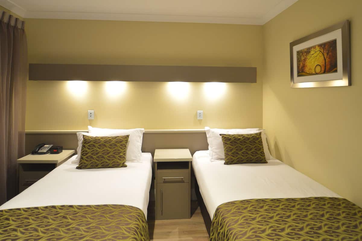 Quarto de hotel com duas camas de solteiro, uma mesa de cabeceira com telefone e um candeeiro fixado na parede.