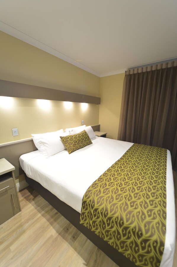 Quarto de hotel com cama, mesa de cabeceira, luminária e janela com cortinas.