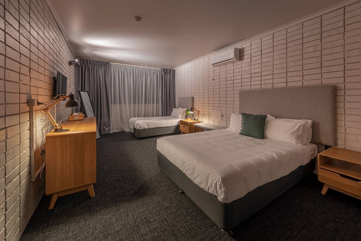 Quarto de hotel com duas camas, uma escrivaninha, uma cadeira, um abajur e uma janela com cortinas.