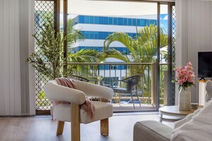 Uma sala de estar moderna com um sofá branco, uma mesa de centro e uma TV. É visível uma varanda com plantas e um edifício através das portas de vidro.