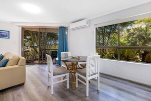 Quarto moderno com mesa de vidro, cadeiras brancas, sofá bege e janelas amplas com vista para o verde.