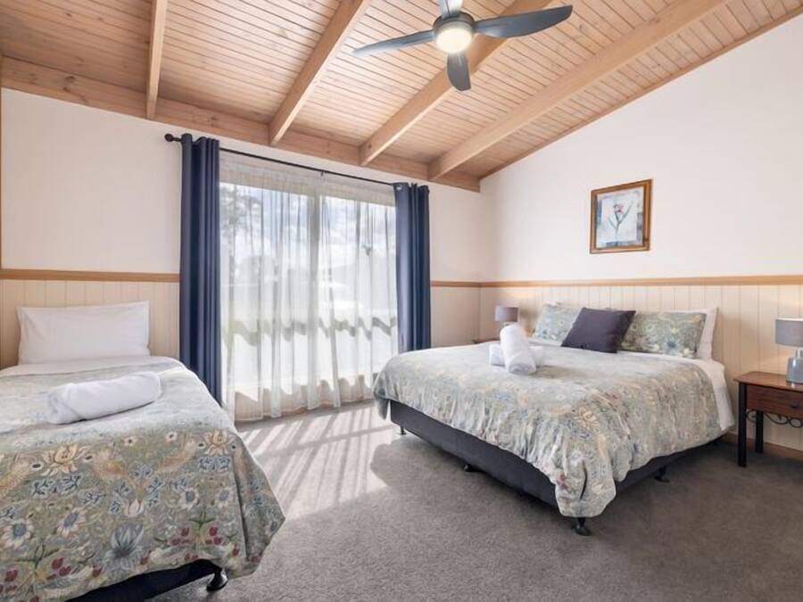 Quarto com duas camas, um ventilador de teto, janela com cortinas e mesa de cabeceira com abajur.