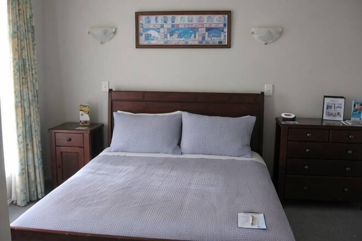 Uma cama bem arrumada com cabeceira, mesa de cabeceira com luminária, cômoda com gavetas e luminária fixa na parede.