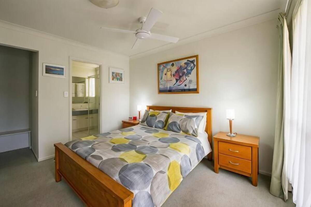 Quarto com cama, mesas de cabeceira, ventoinha no teto, um quadro e porta do banheiro.