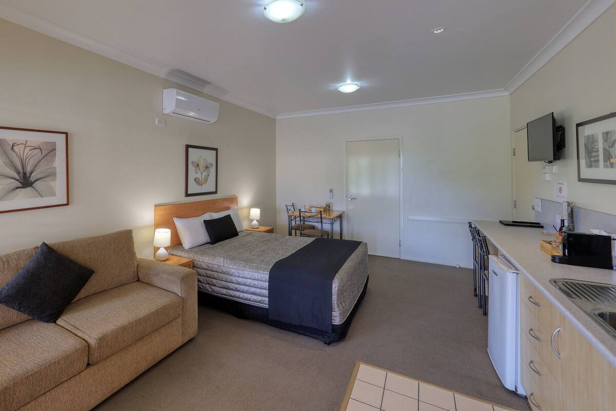 Quarto de hotel com cama, sofá, escrivaninha e cozinha compacta.