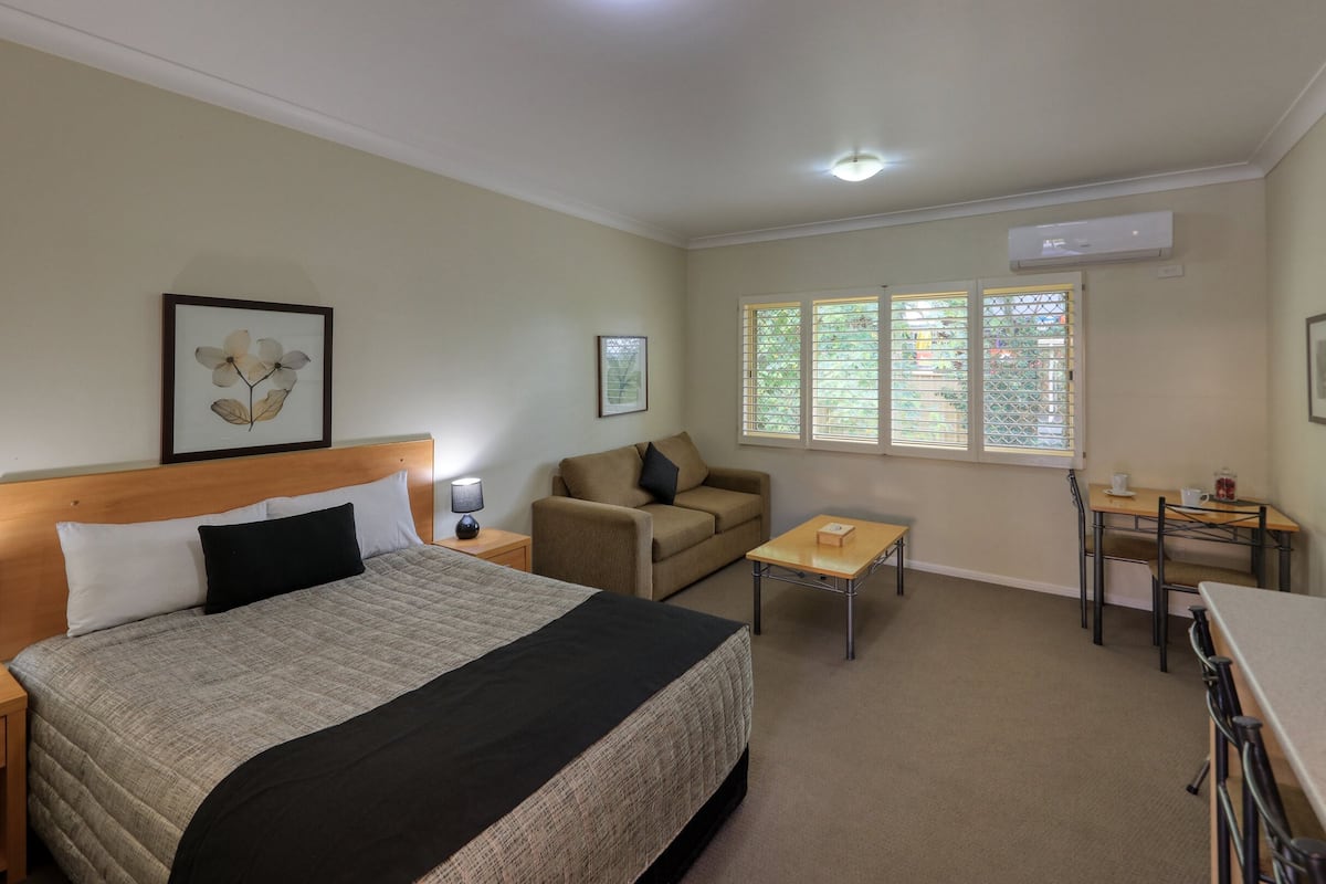 Quarto de hotel com cama, sofá, mesa pequena e escrivaninha. Janelas com persianas, luminária no teto e aparelho de ar condicionado.