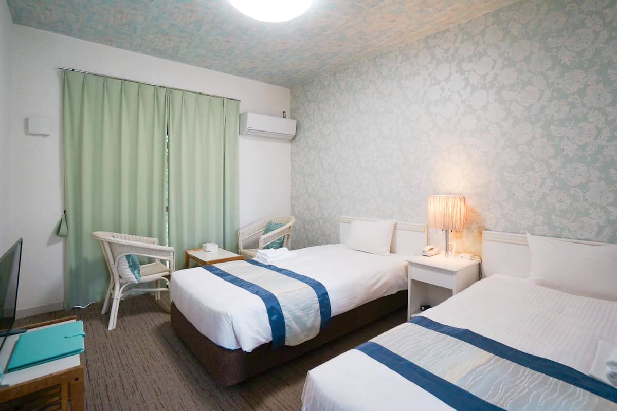 Quarto de hotel com duas camas, uma escrivaninha, uma cadeira e um abajur.