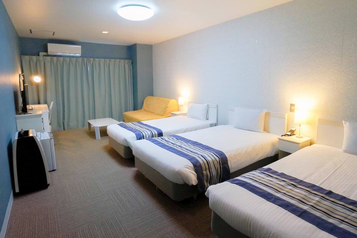 Quarto de hotel com duas camas, uma mesinha, uma cadeira, uma escrivaninha com computador e uma janela com cortinas.