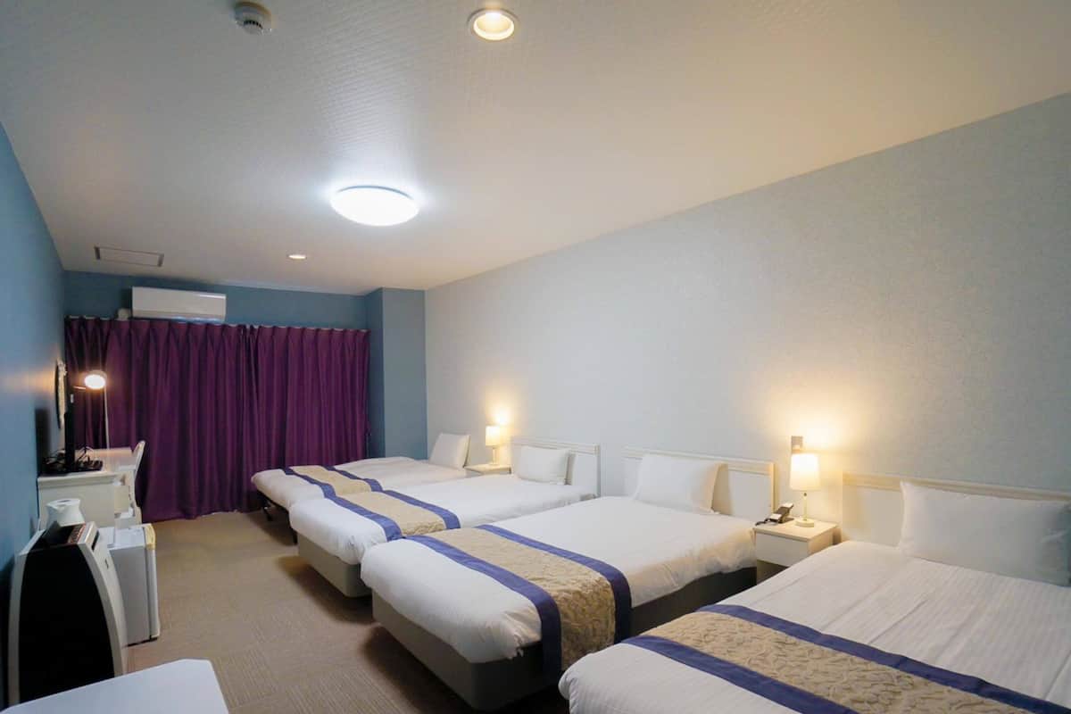 Quarto de hotel com quatro camas de solteiro, uma escrivaninha, uma cadeira, um abajur e um aparelho de ar condicionado.