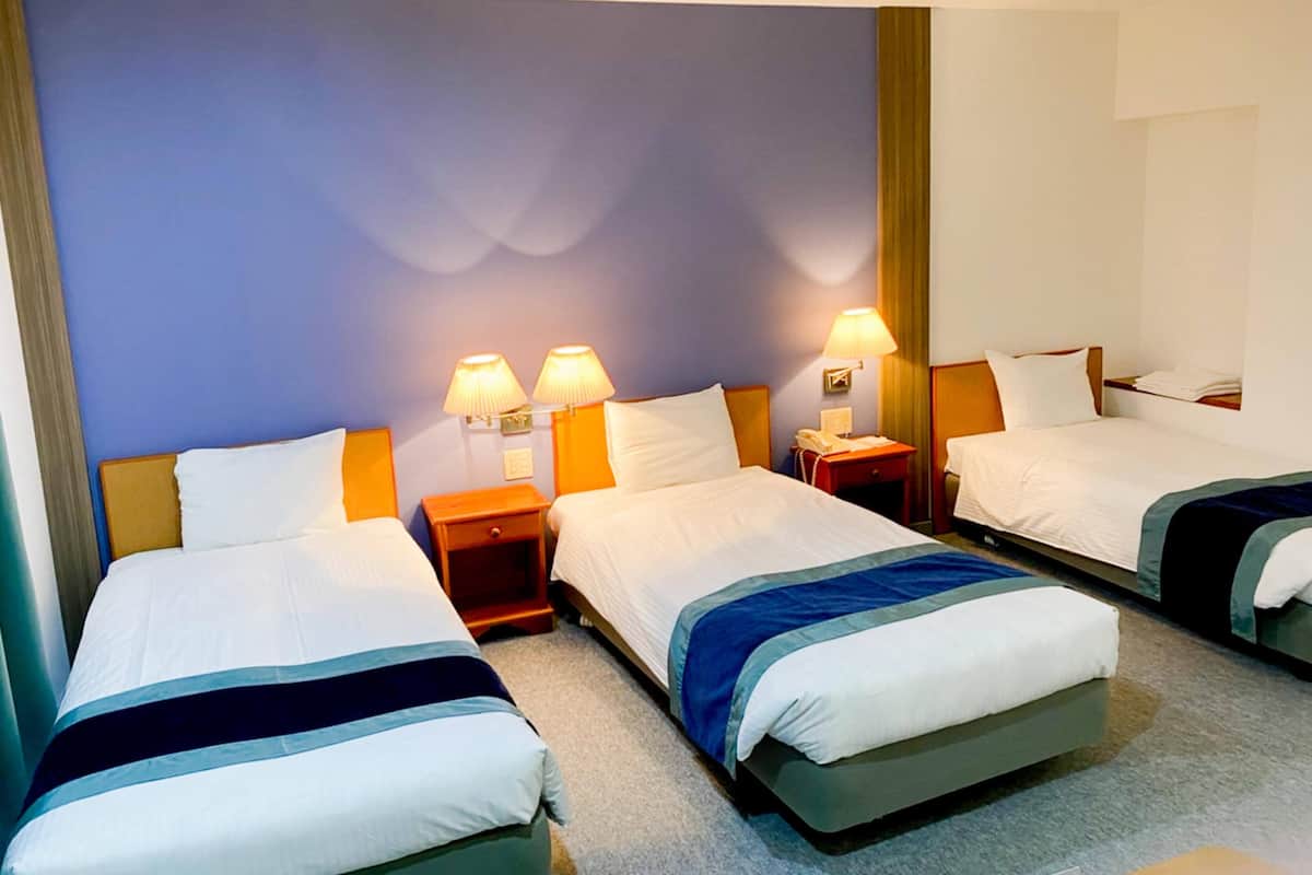 Quarto de hotel com três camas de solteiro, cabeceira de madeira, mesa de cabeceira com luminária e prateleira na parede com um telefone.