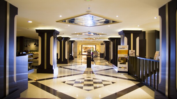 Um hall de hotel com piso xadrez, colunas imponentes e um lustre central.