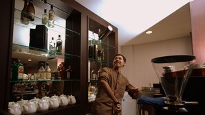 Uma área de bar com prateleiras de garrafas e copos, uma máquina de café e uma pessoa em pé.