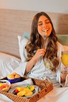 Uma pessoa sentada na cama com uma bandeja de café da manhã contendo croissants, frutas e um copo de suco de laranja.