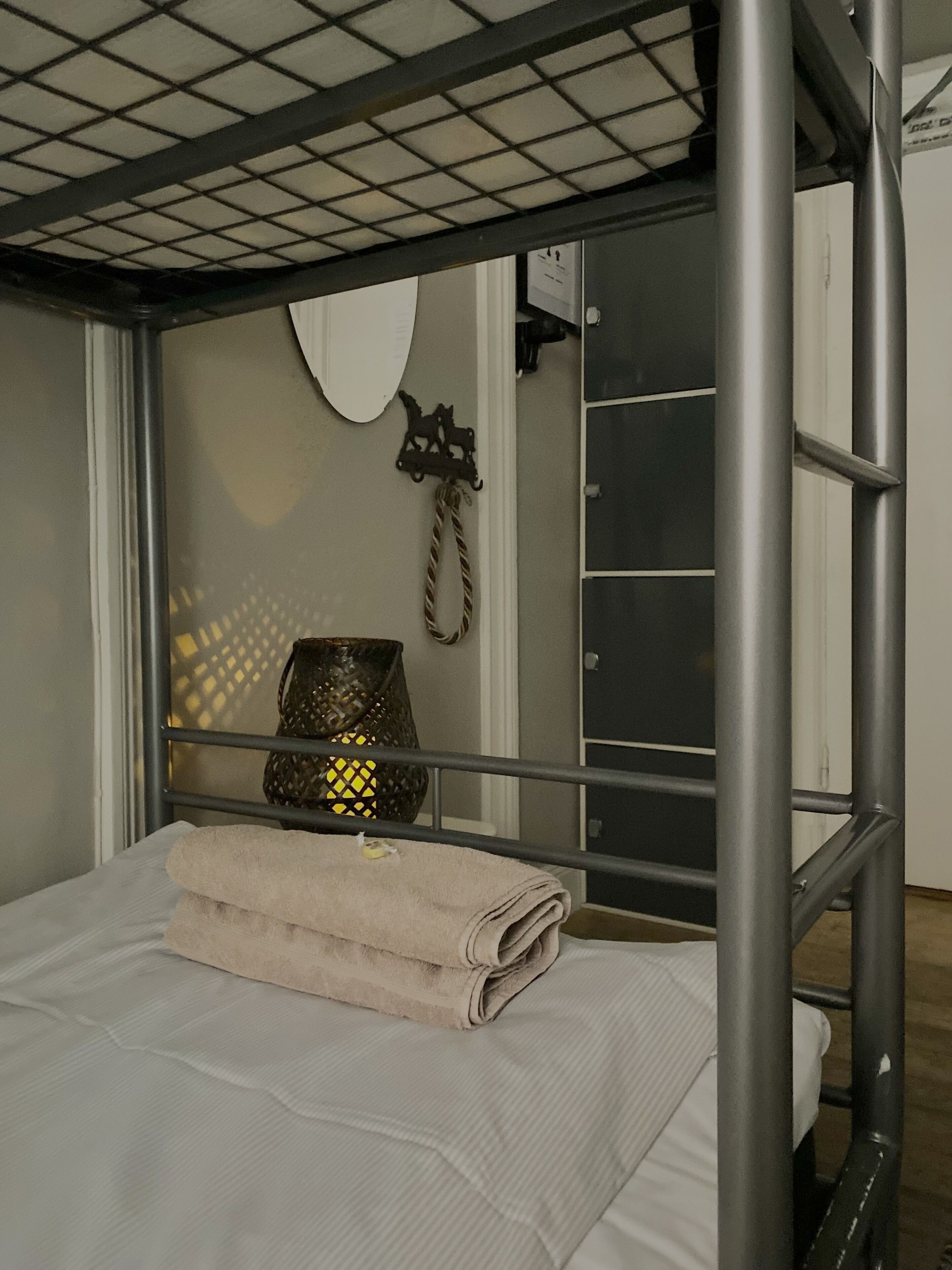 Uma cama beliche com estrutura de metal, um colchão branco e uma toalha enrolada. Há um espelho fixado na parede e um enfeite de parede com um franjário.