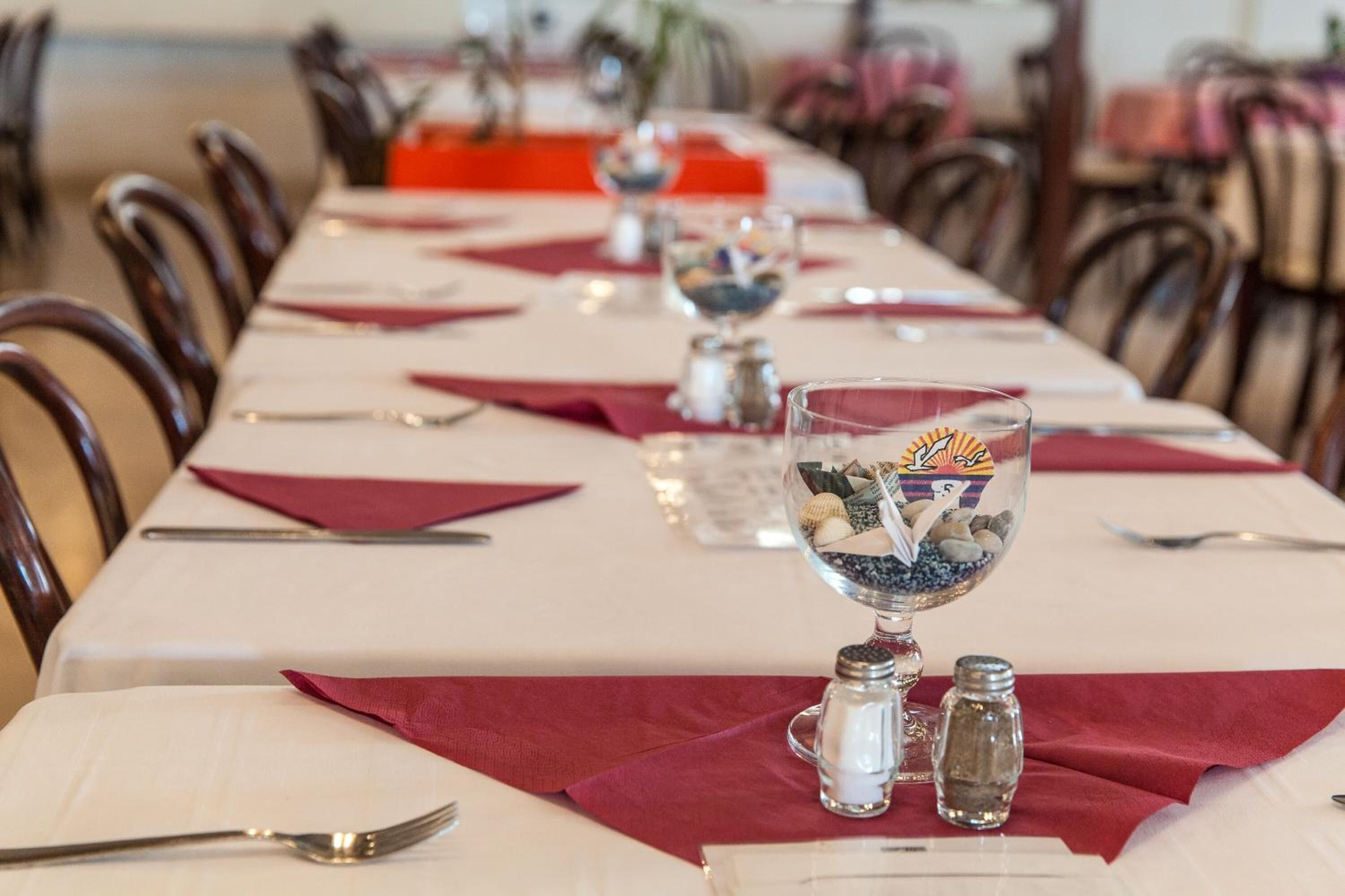Mesa de jantar posta com guardanapos vermelhos, saleiro e pimenteiro, e um centro de mesa de vidro decorativo.