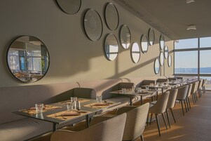 Um restaurante com um balcão comprido, vários espelhos redondos na parede e vista para a cidade através de grandes janelas.