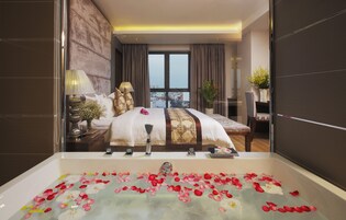 Quarto de hotel moderno com uma cama grande, uma banheira cheia de pétalas de rosa e vista para a cidade pela janela.