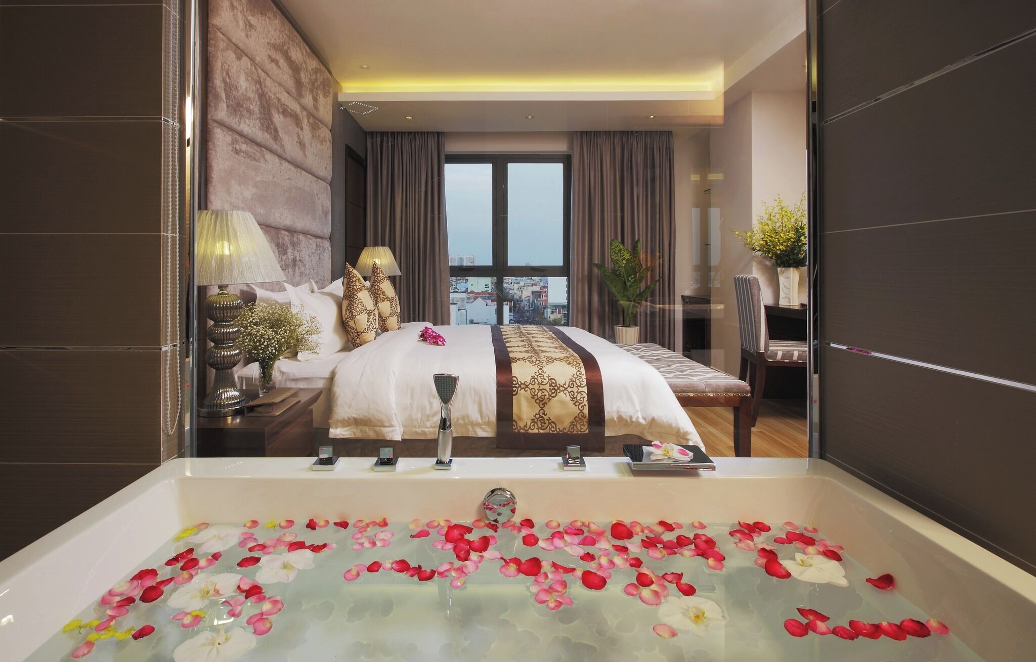 Quarto de hotel com uma cama grande, uma banheira cheia de pétalas de rosa, vista para a cidade e um espaço de refeições com mesa e cadeiras.