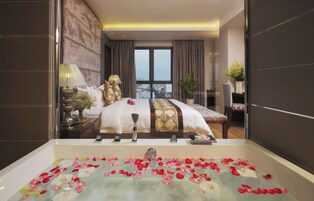 Quarto de hotel com uma cama grande, uma banheira cheia de pétalas de rosa, vista para a cidade e um espaço de refeições com mesa e cadeiras.
