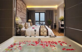 Quarto de hotel com uma cama grande, banheiro com banheira cheia de pétalas de rosa e vista para a cidade pela janela.