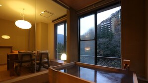 Uma onsen japonesa tradicional com banheira de madeira, mesa com cadeiras e grandes janelas com vista para um morro arborizado.