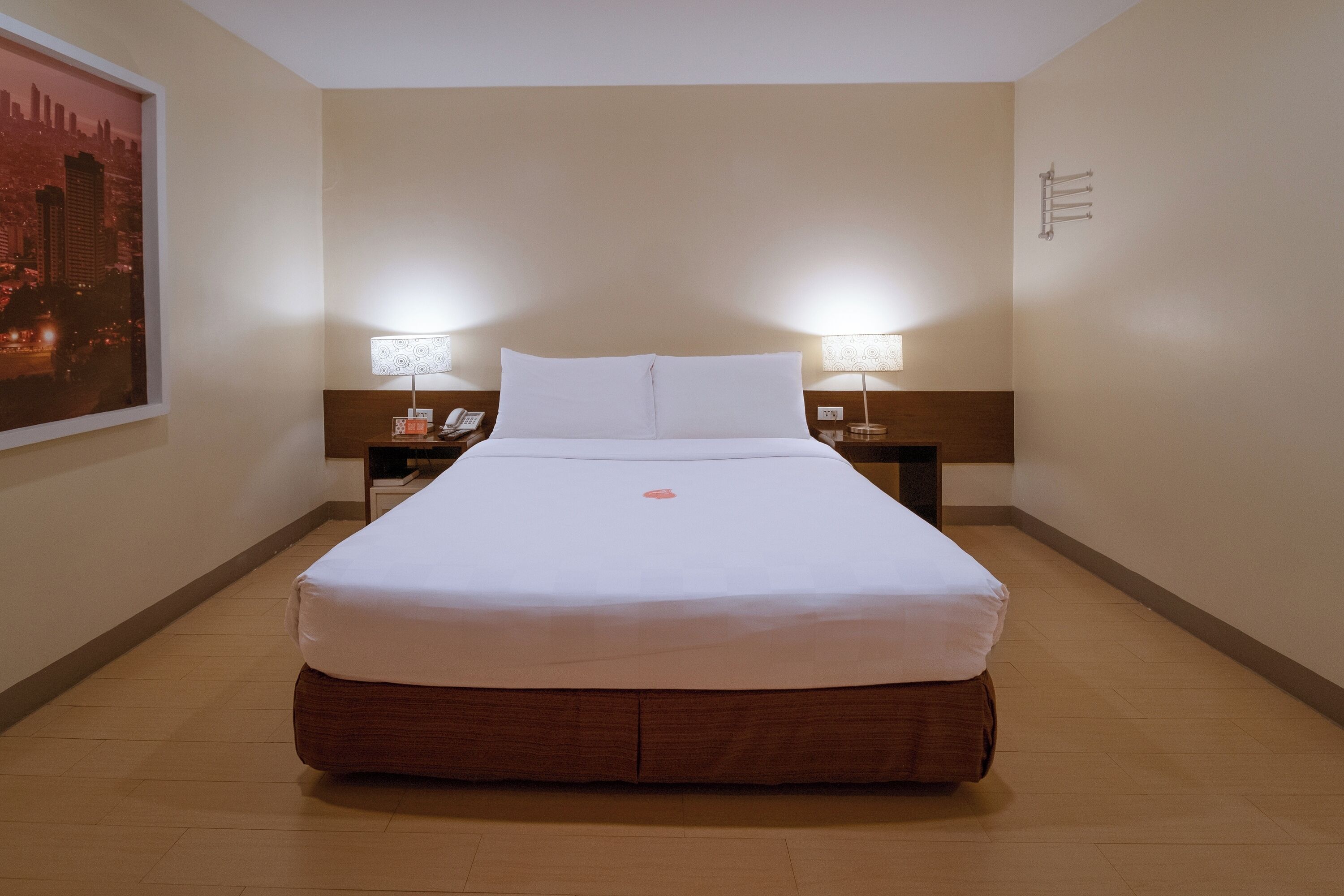Cama bem arrumada em quarto de hotel com vista para a cidade, duas luminárias de cabeceira e mesa de cabeceira com telefone e uma mesinha.