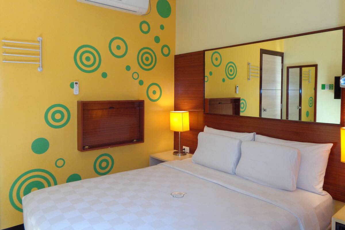Quarto de hotel com uma cama, mesa de cabeceira, abajur, espelho e parede com desenhos circulares.