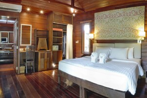 Quarto com piso de madeira, uma cama grande, um guarda-roupa, uma escrivaninha com cadeira e um banheiro com espelho e pia.