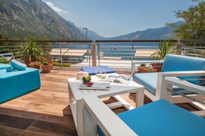 Um terraço com mobiliário azul e branco, uma mesa com uma bandeja de frutas e copos, e vista para o mar e montanhas.