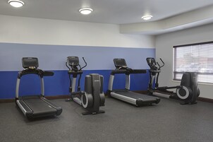Uma academia com quatro esteiras e um aparelho elíptico, uma janela com persianas e uma parede azul e branca.