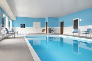Piscina com água azul cristalina, rodeada por paredes azuis e chão de azulejos brancos. Há espreguiçadeiras e um vestiário com chuveiro.