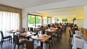 Uma ampla sala de jantar com várias mesas e cadeiras, grandes janelas com cortinas e vista para o verde lá fora.