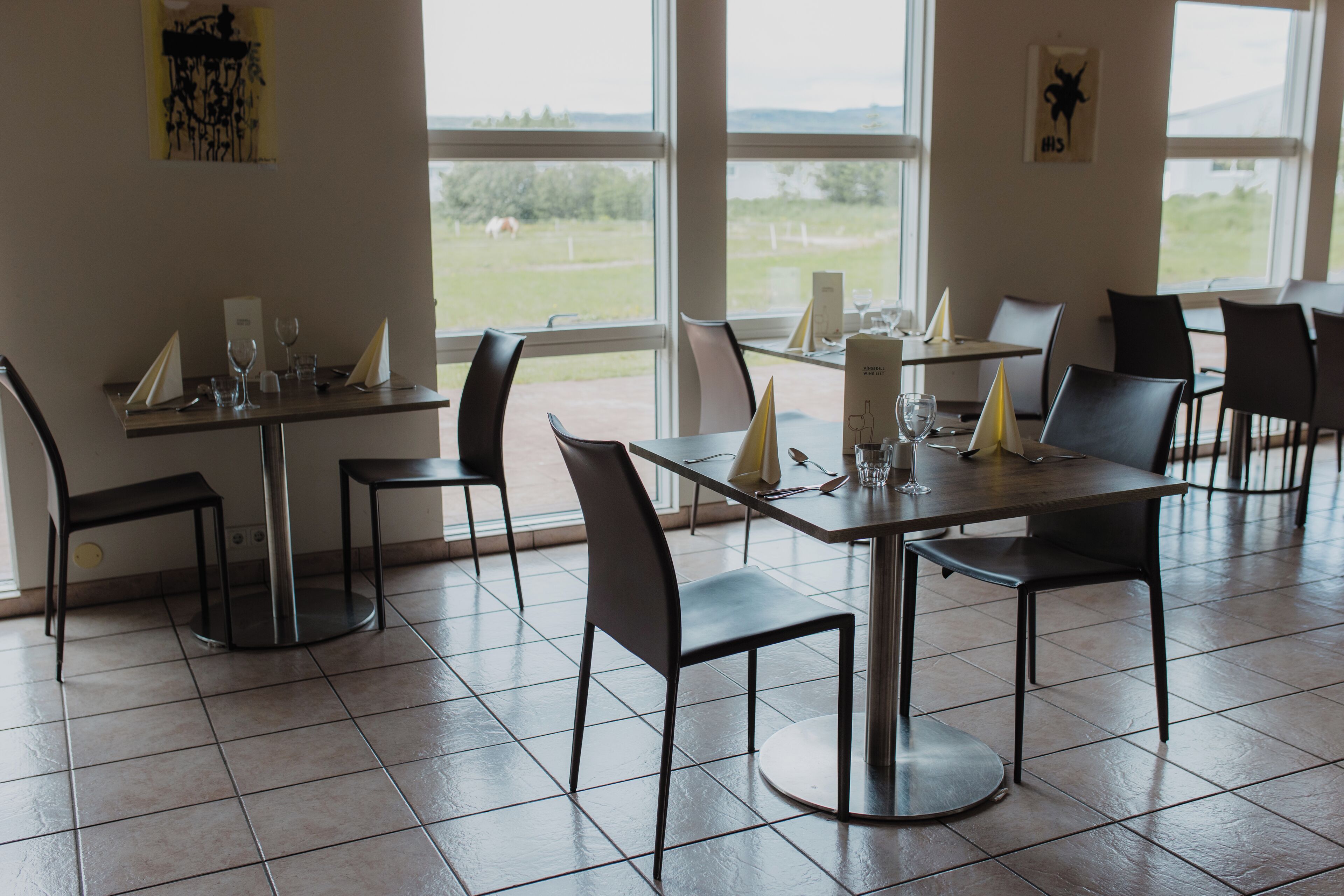 Um restaurante com decoração moderna, com uma mistura de mesas redondas e retangulares, cadeiras pretas e vista para o exterior através de grandes janelas.