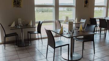 Um restaurante com decoração moderna, com uma mistura de mesas redondas e retangulares, cadeiras pretas e vista para o exterior através de grandes janelas.