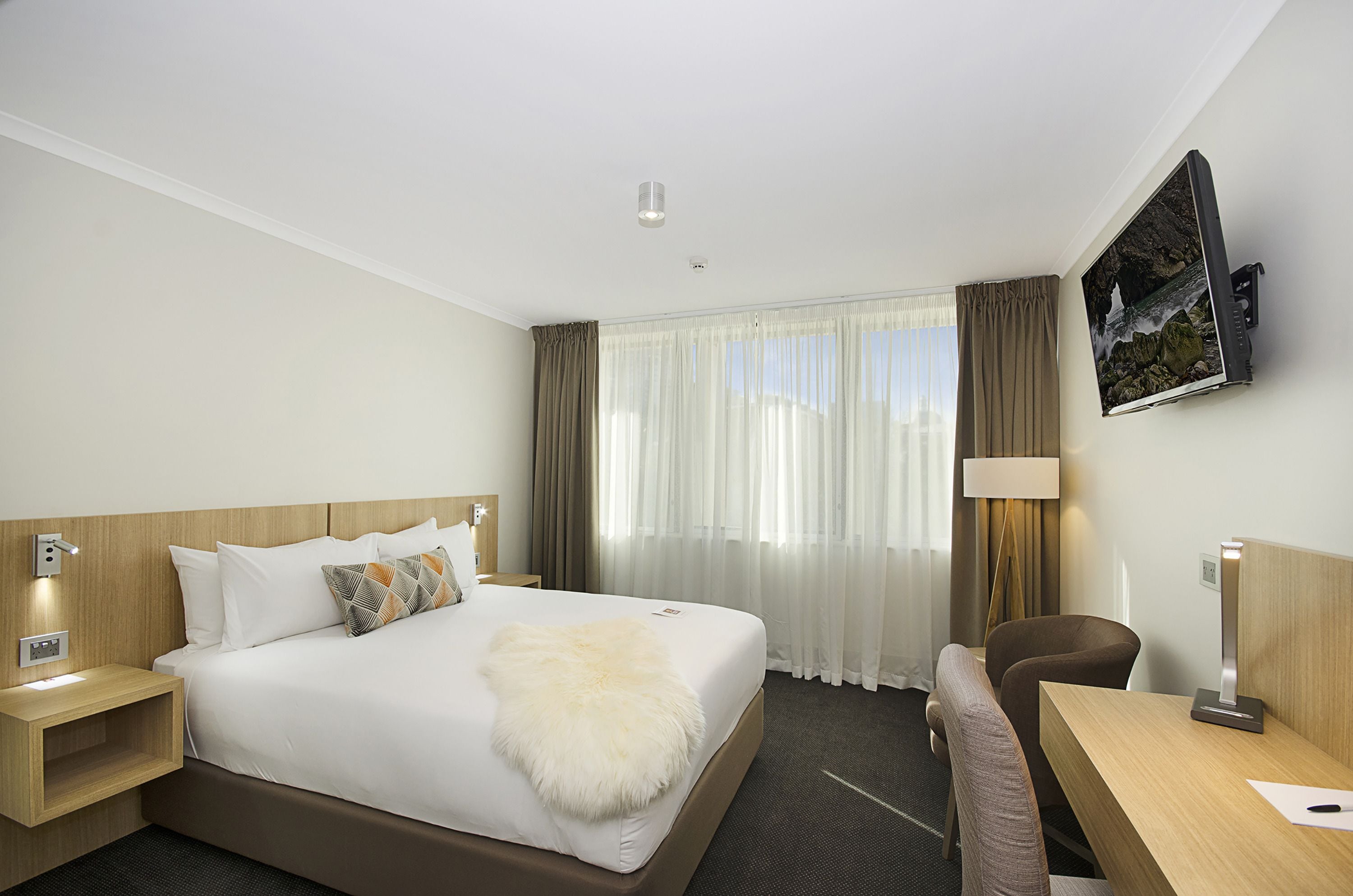 Quarto de hotel com uma cama grande, uma escrivaninha, uma cadeira, uma televisão na parede e uma janela com cortinas.
