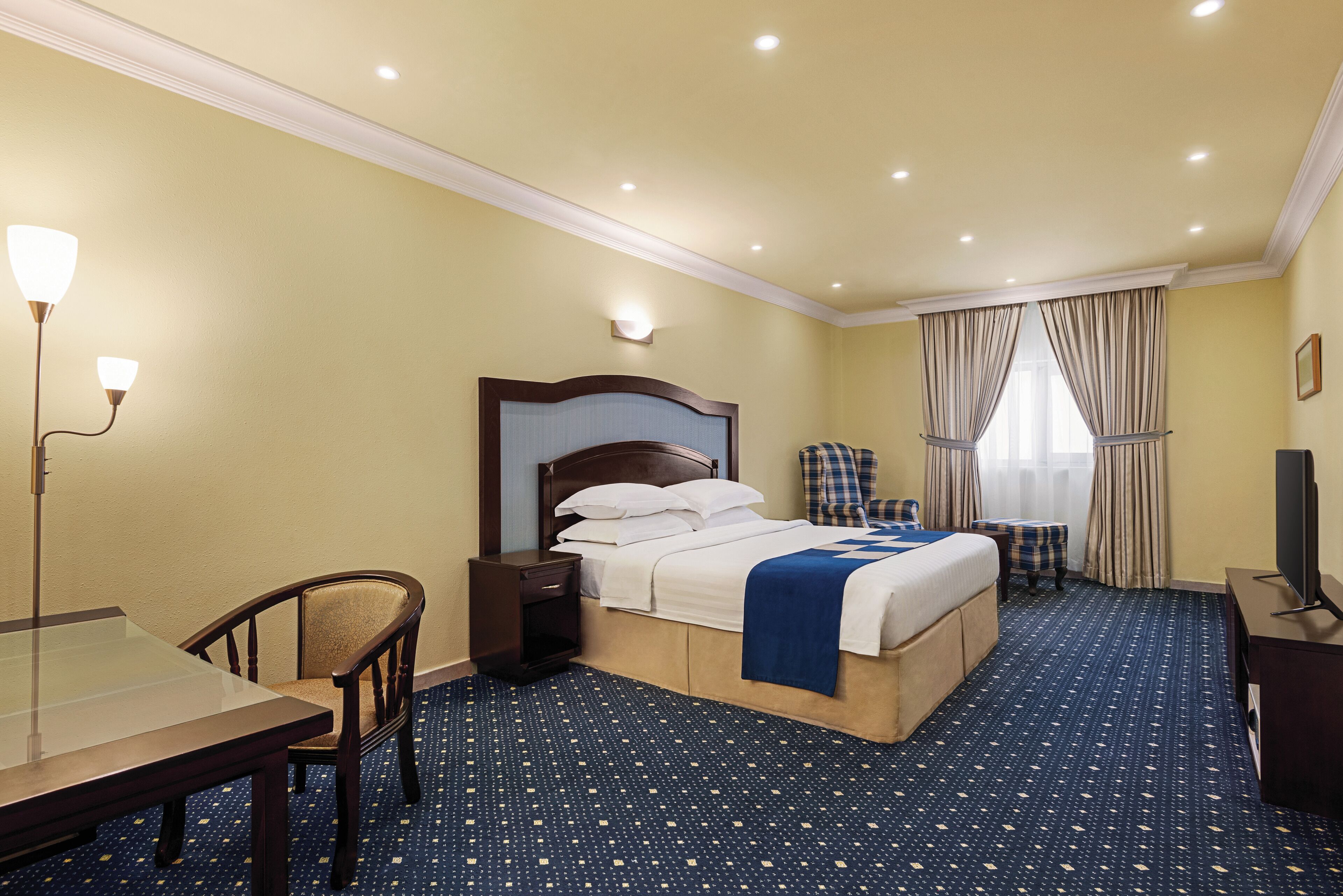 Quarto de hotel com cama, escrivaninha, cadeira, televisão e janela com cortinas.