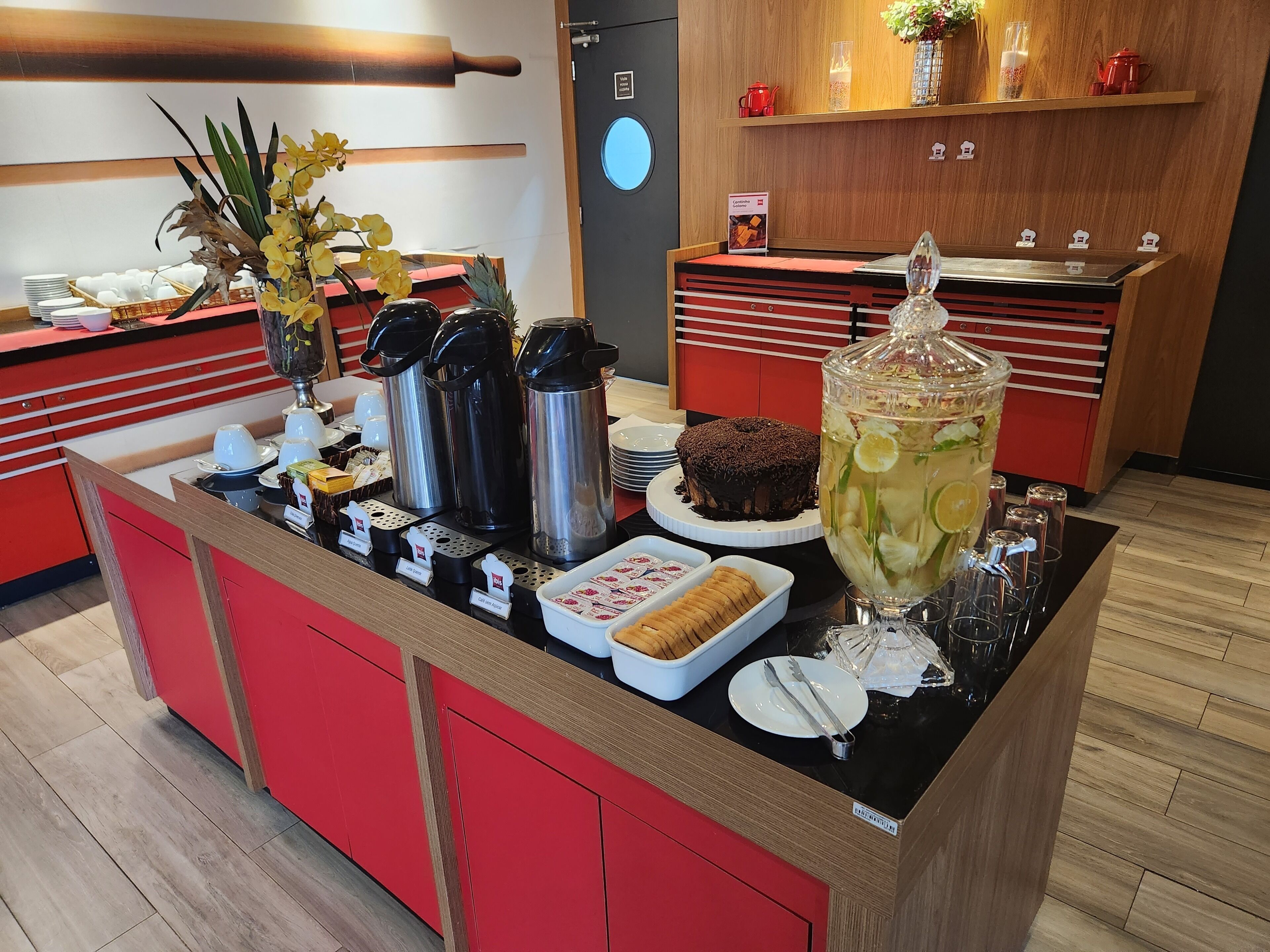 Balcão de buffet com diversos alimentos, máquinas de café e um vaso decorativo.