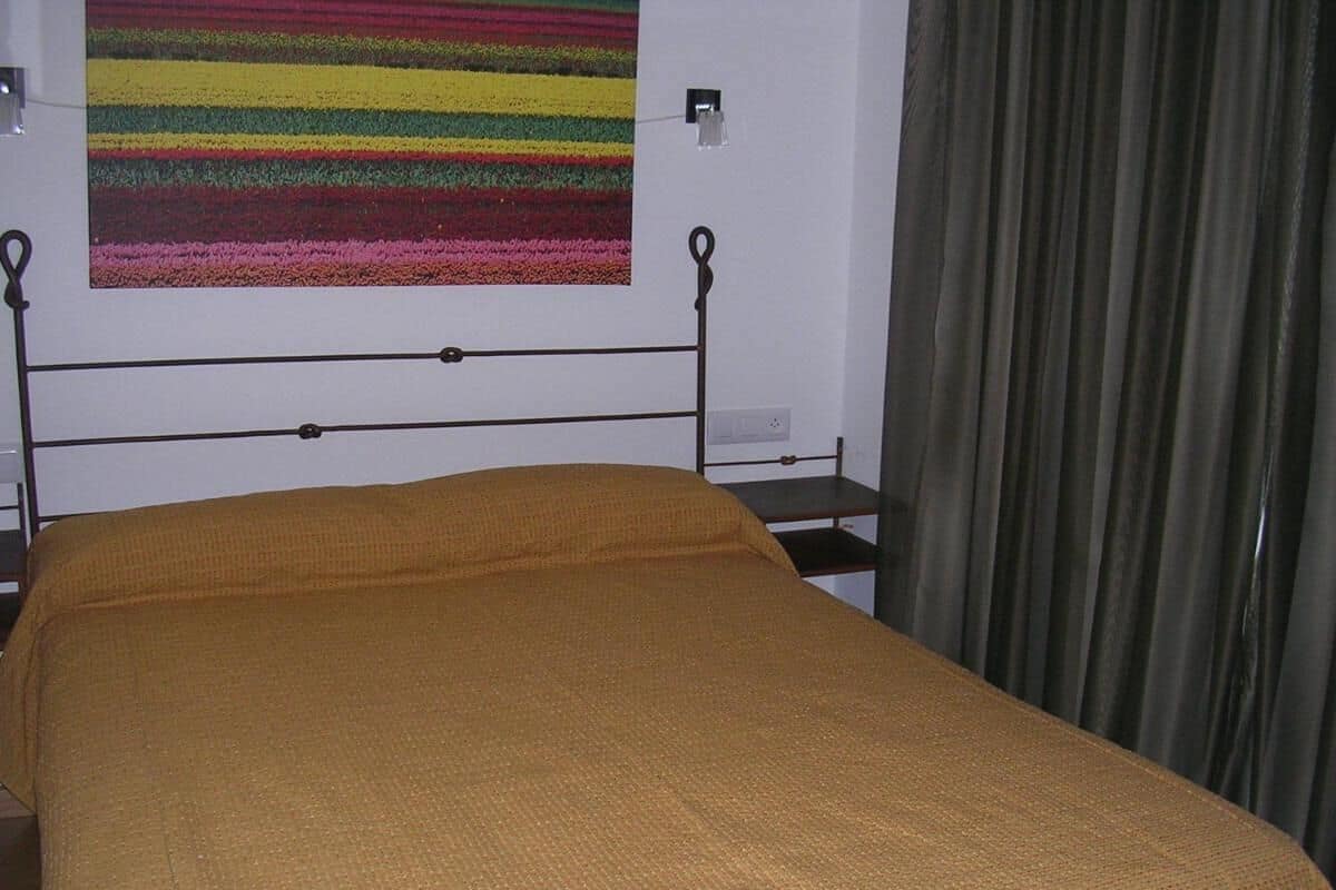 Uma cama de solteiro com um cobertor amarelo-mostarda em um quarto com arte de parede listrada e uma cortina.