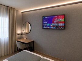 Quarto de hotel com televisão de tela plana fixada na parede exibindo um canal esportivo, um espelho redondo, uma escrivaninha com cadeira e uma cama com cabeceira cinza.