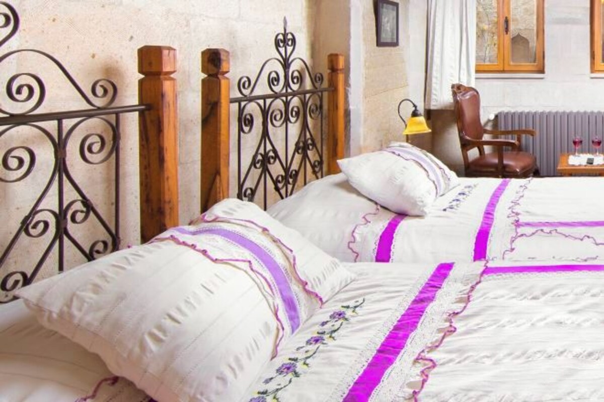 Cama com cabeceira de metal, pilares de madeira e colcha branca com detalhes em roxo.