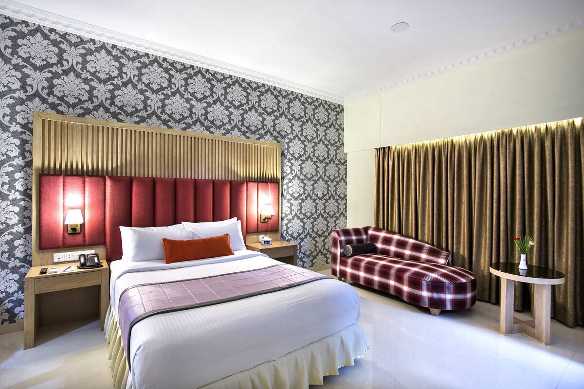 Quarto de hotel com uma cama grande, cabeceira estofada vermelha, sofá vermelho e branco xadrez, mesa lateral redonda e uma cortina.