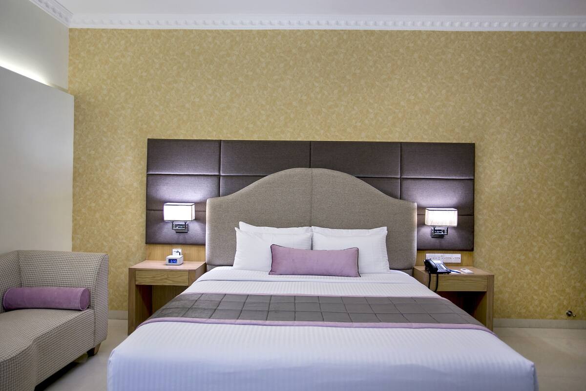 Quarto de hotel com uma cama grande, duas mesas de cabeceira, uma cadeira e cabeceira com iluminação embutida.