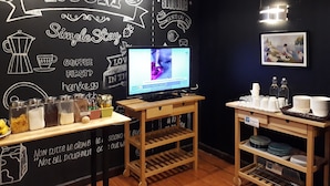 Um quarto acolhedor com uma parede de lousa com várias frases e desenhos, uma televisão exibindo uma imagem roxa e prateleiras com utensílios de cozinha e objetos decorativos.