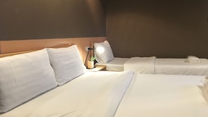 Quarto de hotel com duas camas, cada uma com roupa de cama e almofadas brancas, uma mesinha de cabeceira com abajur e um pequeno vaso de planta.