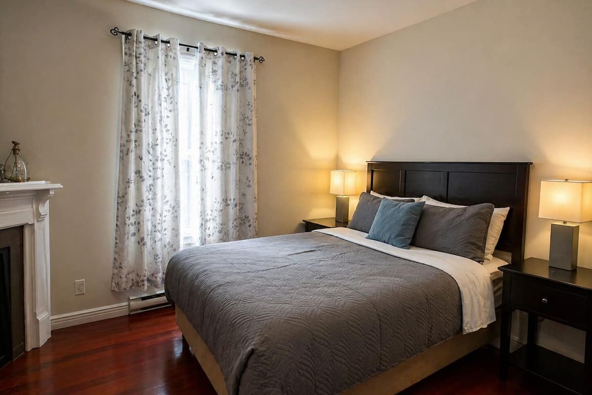 Quarto com cama, duas mesas de cabeceira com abajures, lareira e janela com cortinas florais.